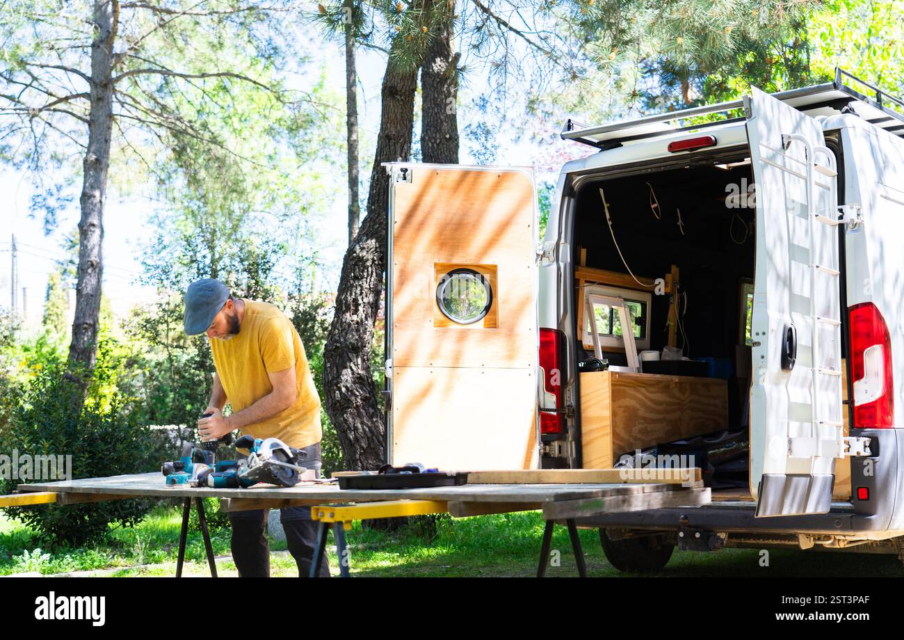 Carpenter working on a camper van conversion project outdoors, using power tools and surrounded ...