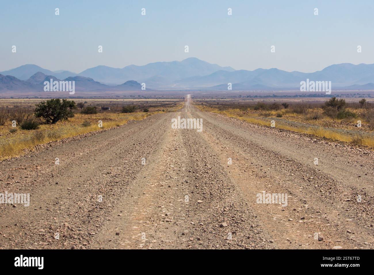 A deserted dirt road leading through the barren Namibian landscape with mountains rising blue in ...