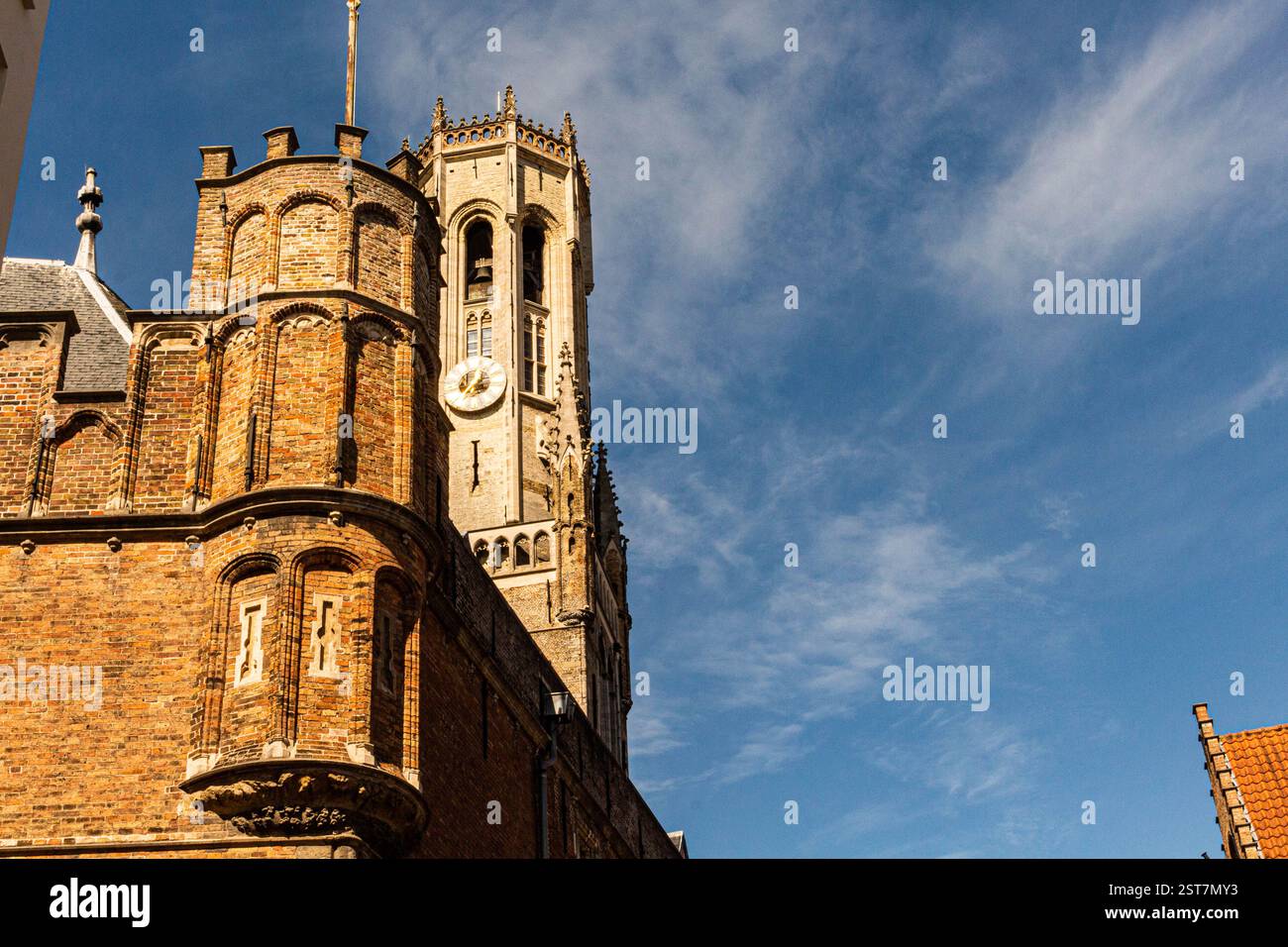 The Belfry of Bruges towers above an old rooftop, framed by a blue sky ...