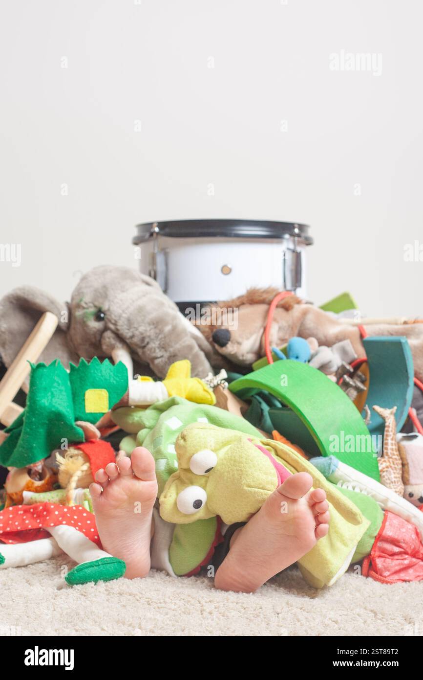 A pile of toys and stuffed animals. Underneath are the feet of a child. They are in a playroom ...