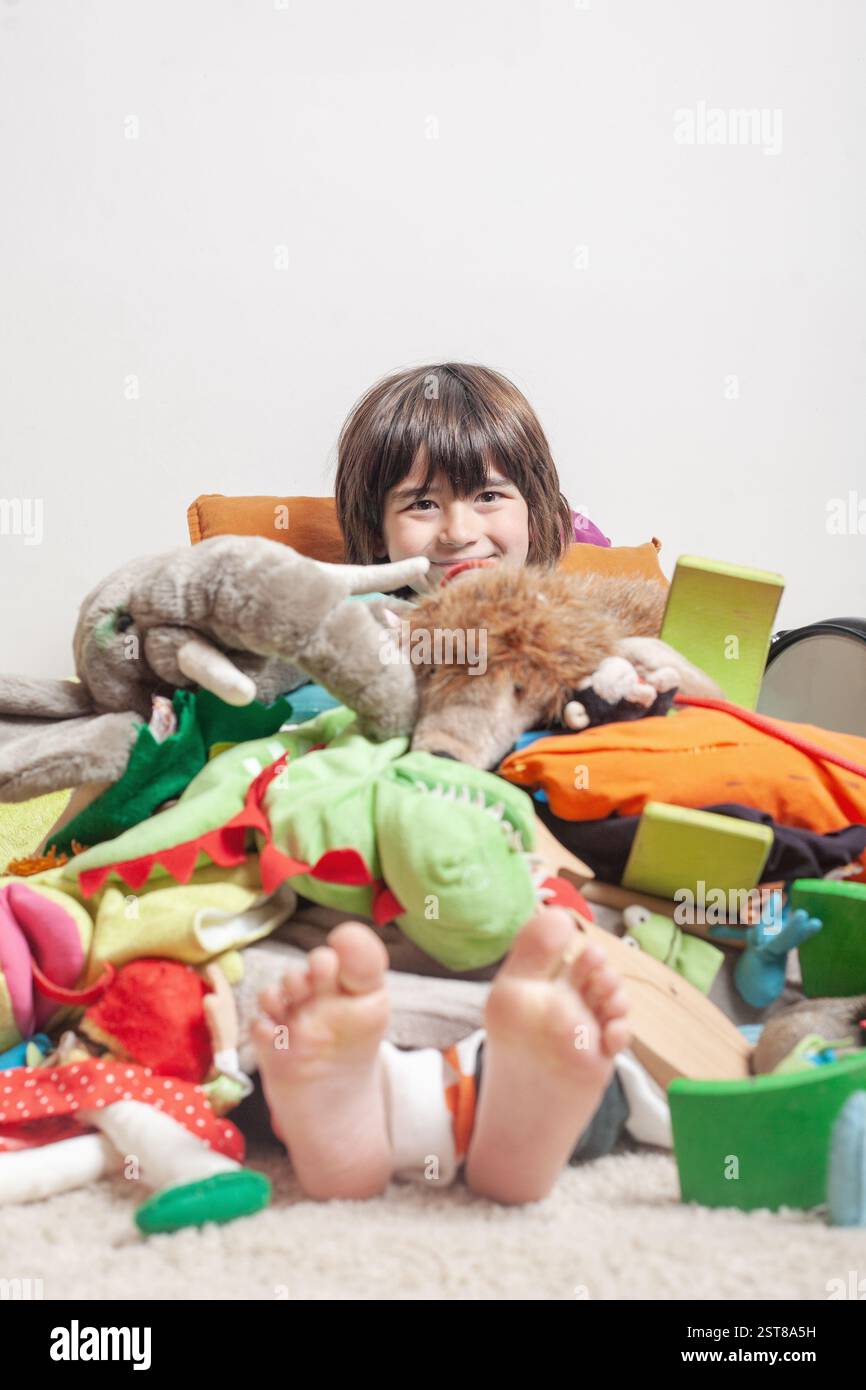 A pile of toys and stuffed animals. Underneath are the feet of a child. They are in a playroom ...