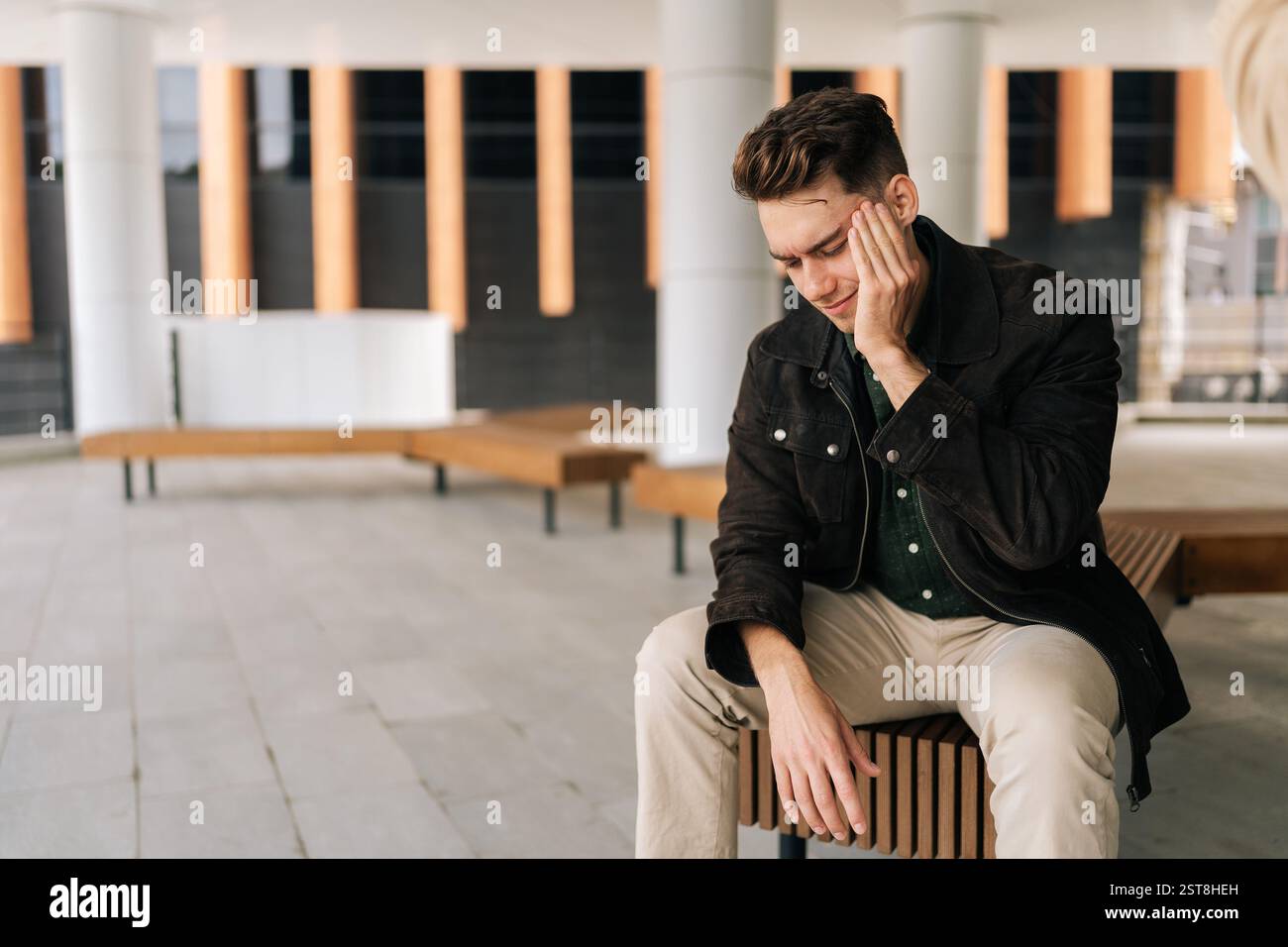 Frustrated sick ill young man touching cheek sitting on urban bench ...
