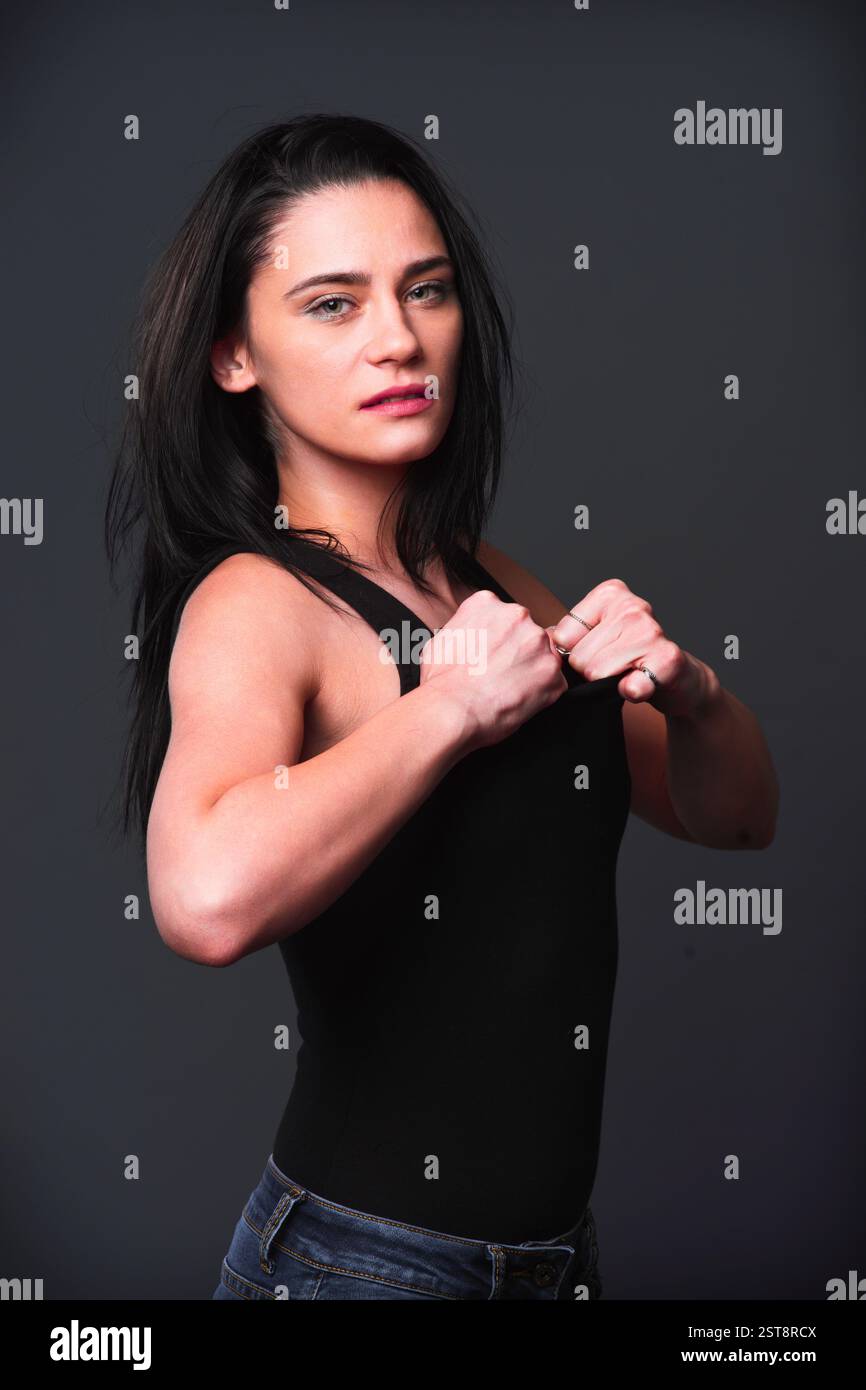 A woman poses confidently, adjusting her black tank top while standing against a neutral ...