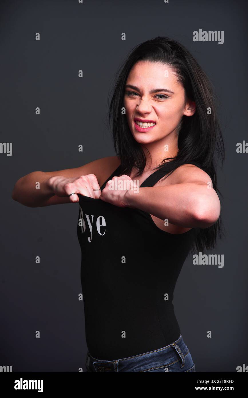 A young woman stands in a studio, showcasing her confidence by tearing open her shirt. Her ...