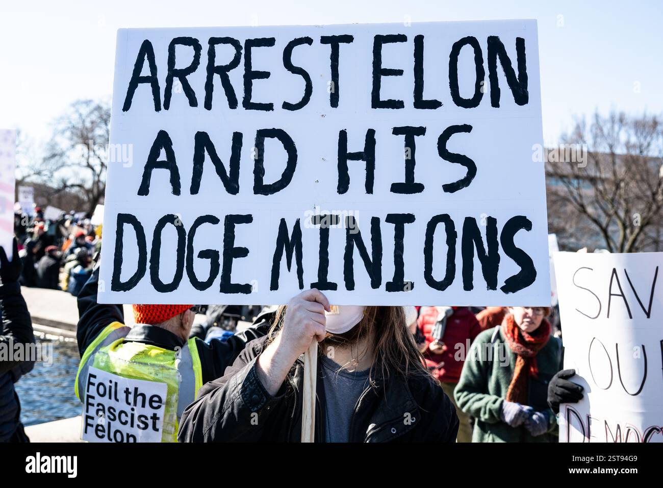 A person with a sign saying "Arrest Elon and his Doge minions" near the ...