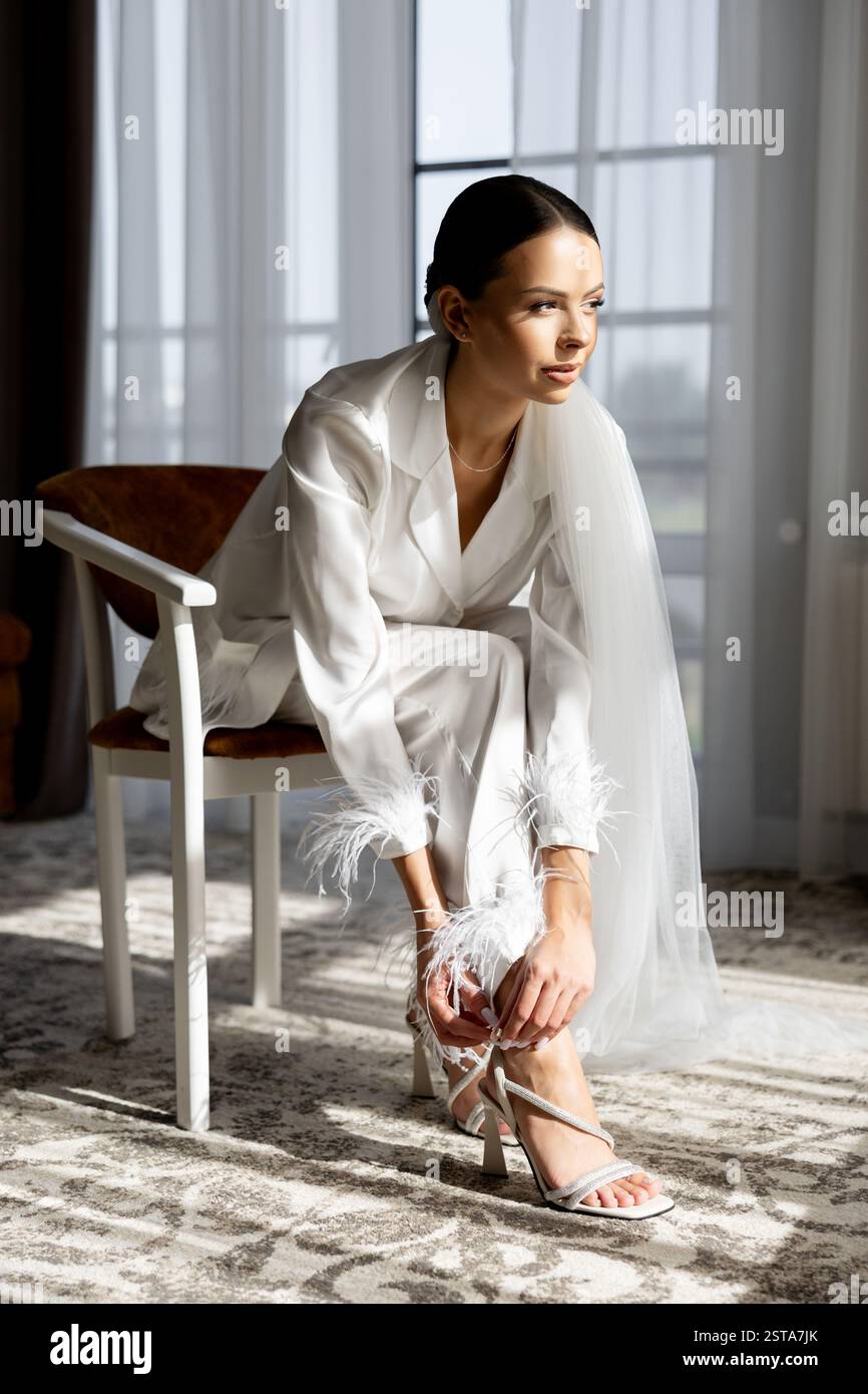 Elegant bride tying her high heels while wearing a white suit and veil ...