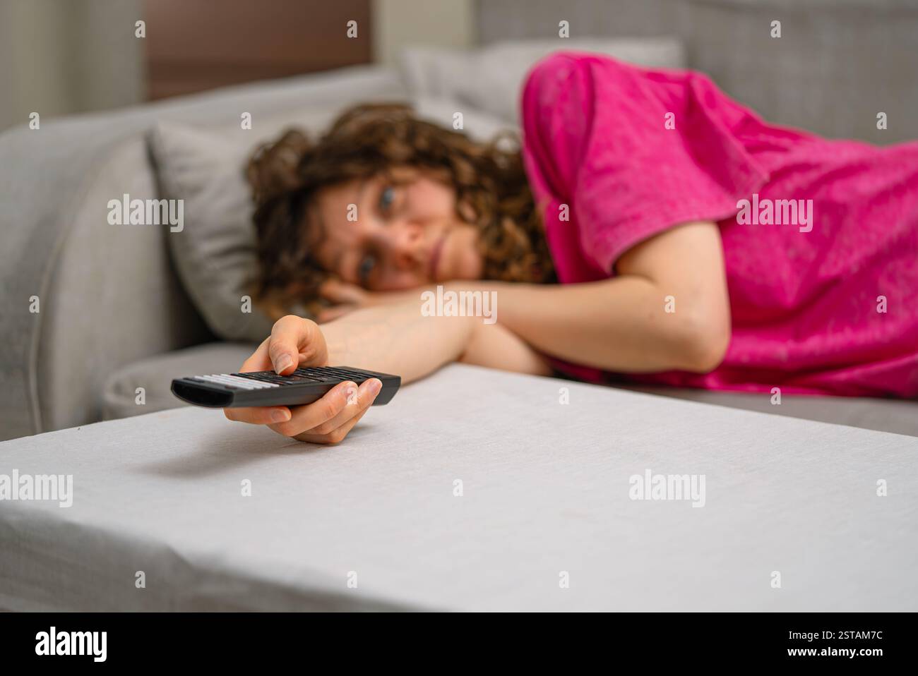 Bored young woman lounging on the gray sofa, flipping through channels ...