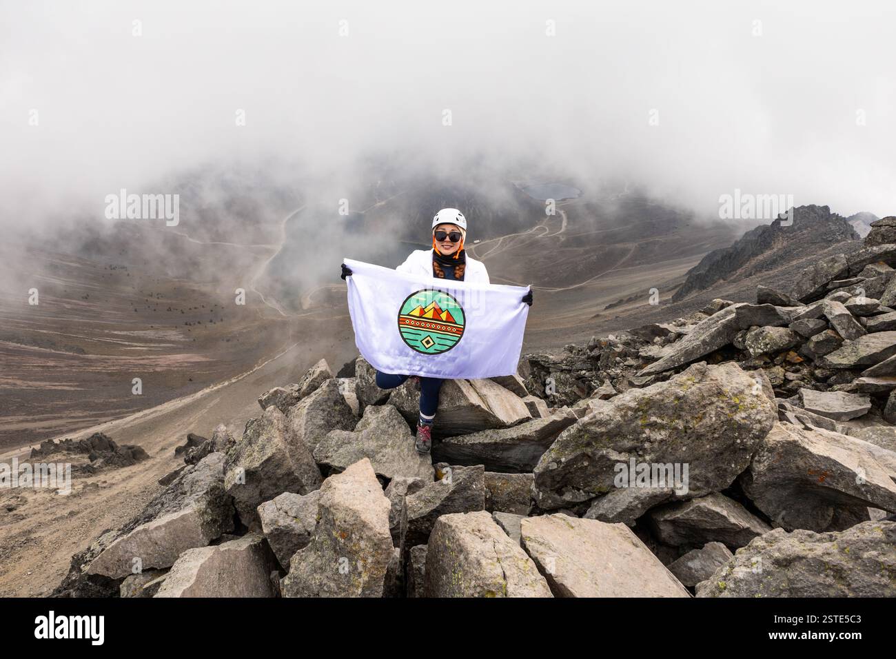 A climber stands atop a rugged volcanic summit, holding a flag while ...