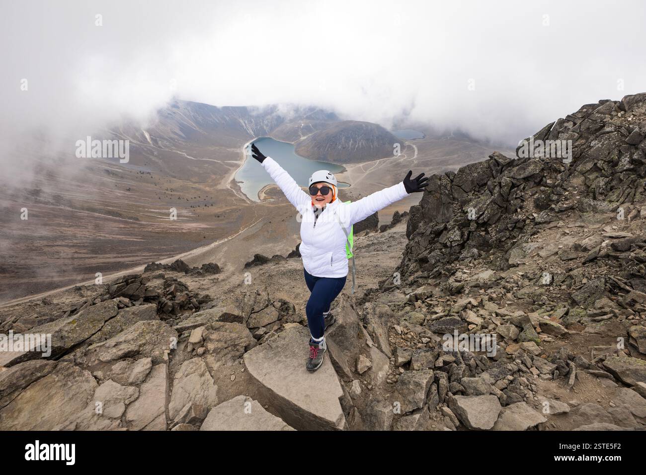 A smiling hiker celebrates at the summit, arms raised in triumph, with ...