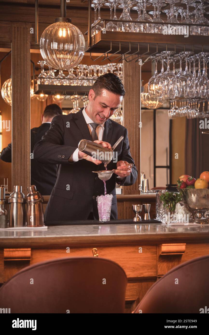 Happy male bartender in black suit pouring cocktail from shaker through ...