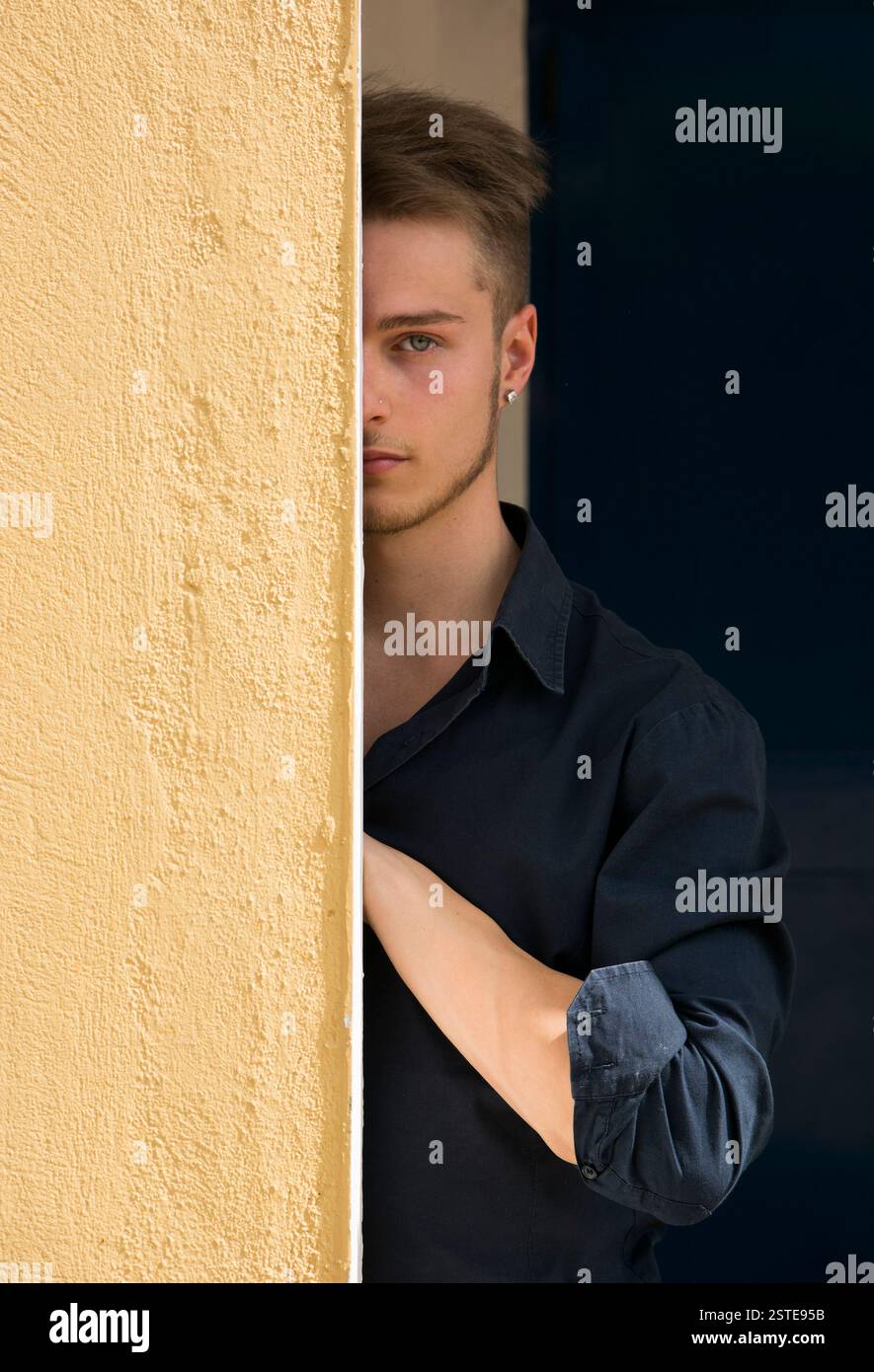 Handsome blond young man hiding half face behind wall looking at camera ...