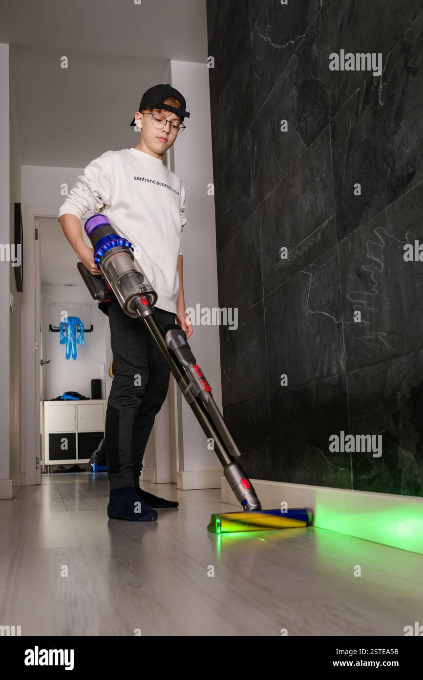 A young individual wearing glasses and a baseball cap vacuums the floor ...
