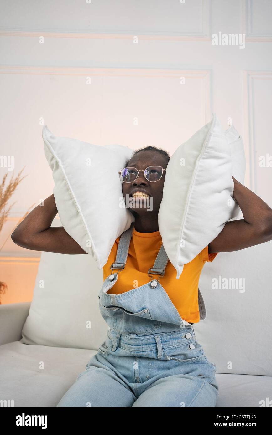 Young black woman using pillows to block out noise, sitting on a couch ...