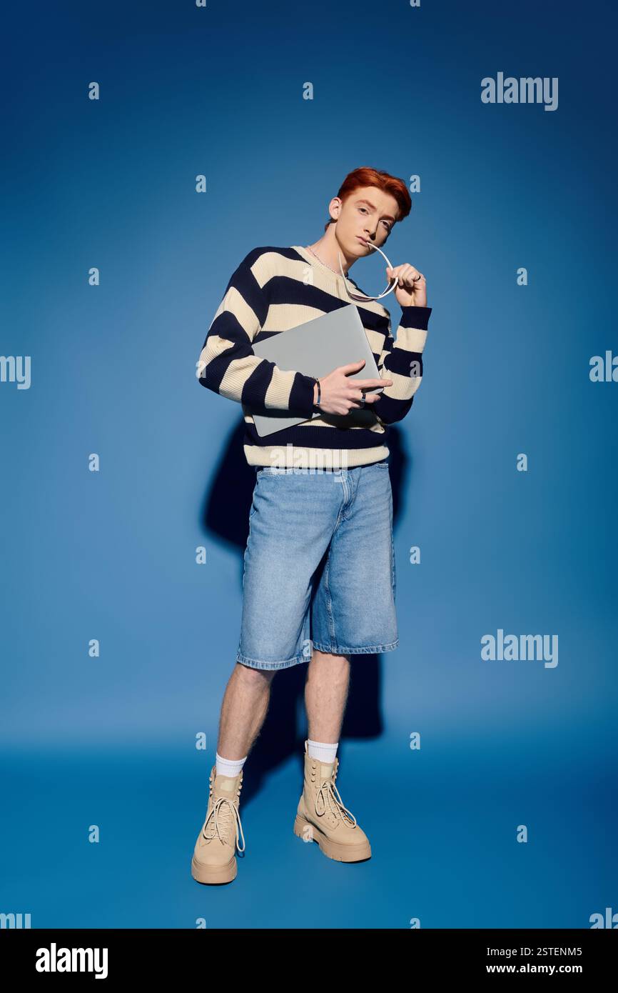 A charming young man in a trendy outfit holds a laptop against a blue ...