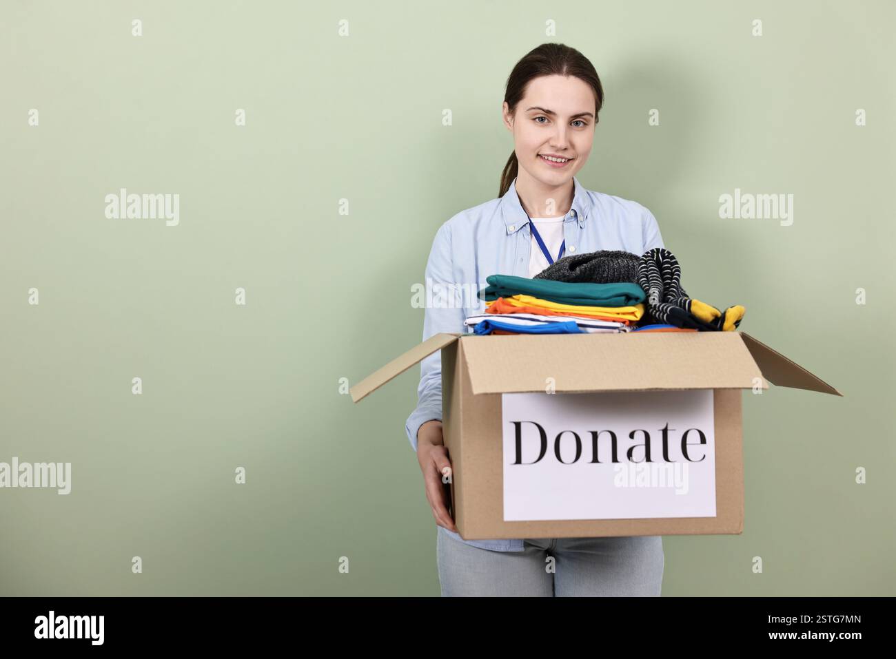 Woman holding donation box with clothes on pale olive background, space ...