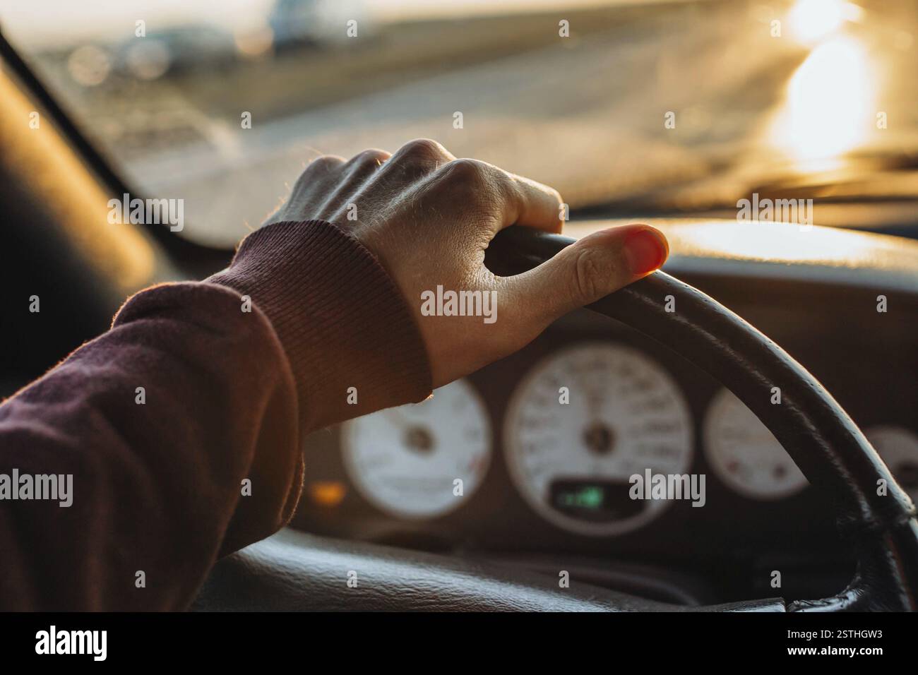 A hand on a steering wheel inside a car with a sunset view outside ...
