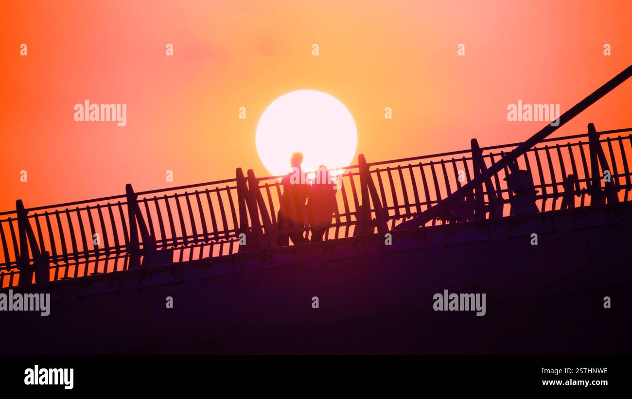 Tourists silhouetted against a vibrant orange sunset sky, crossing the ...