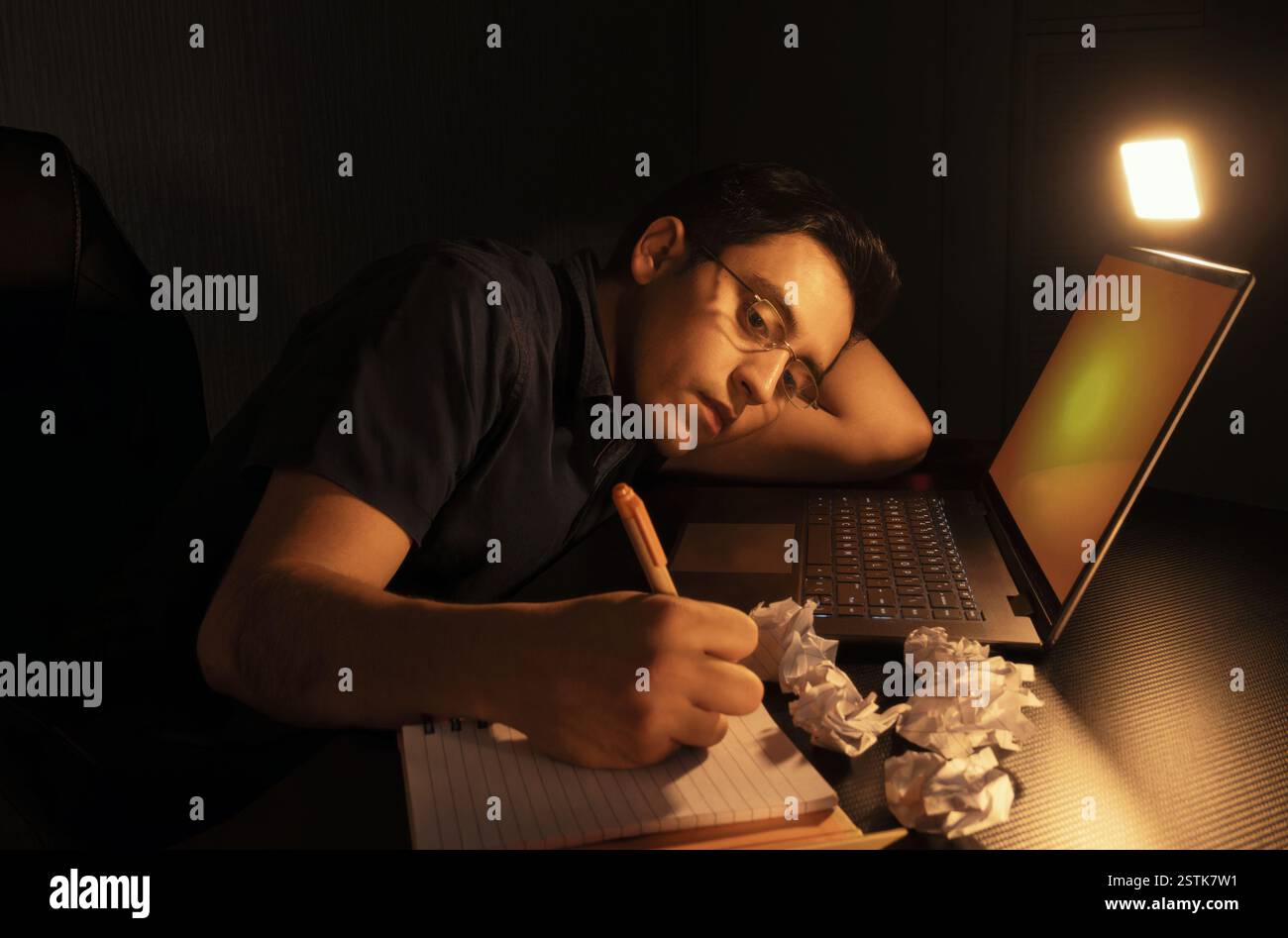 A young man in glasses and casual attire leans over a notebook, writing ...