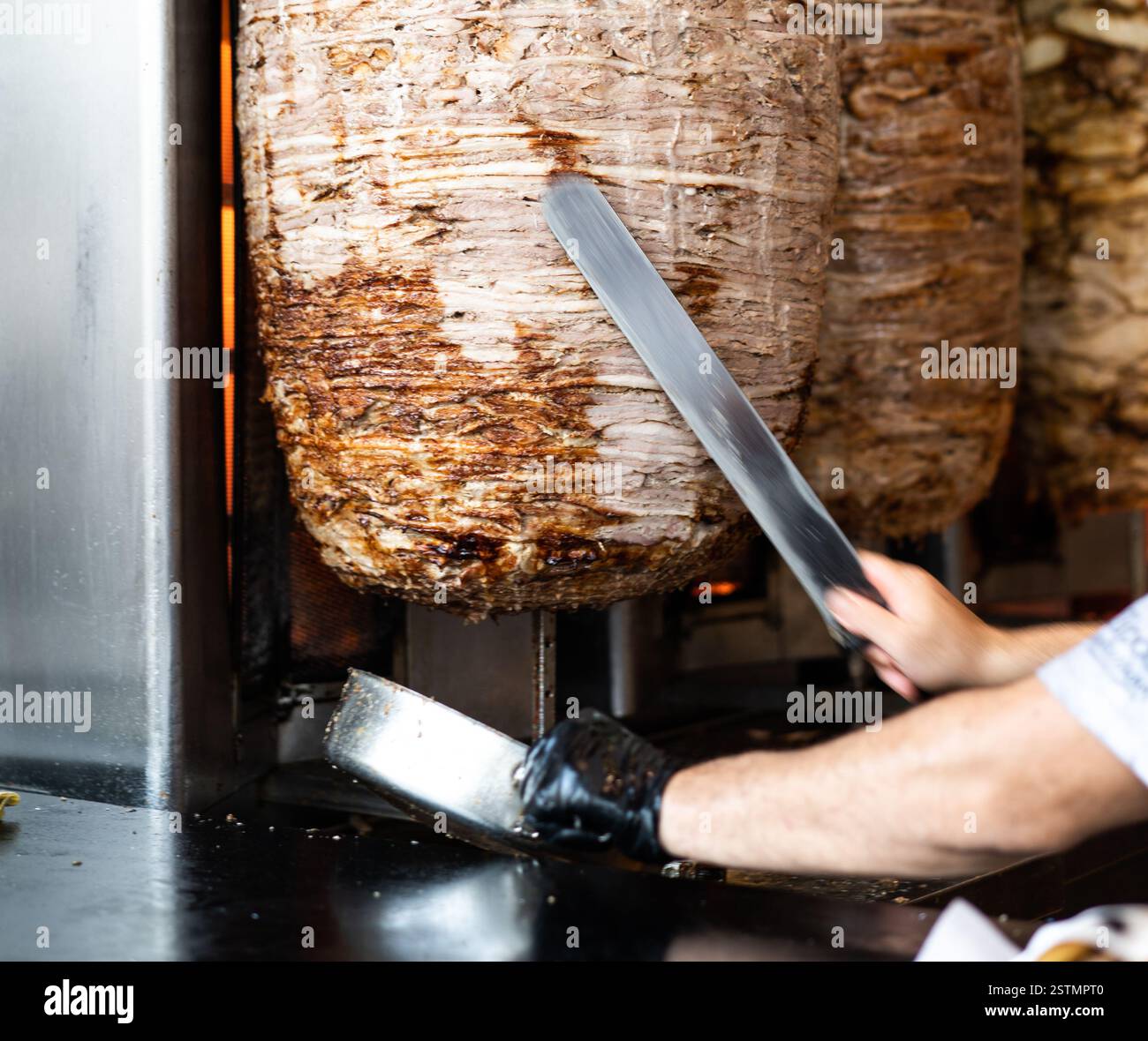 Man's hands holding a knife, cutting slices from a vertical skewer ...