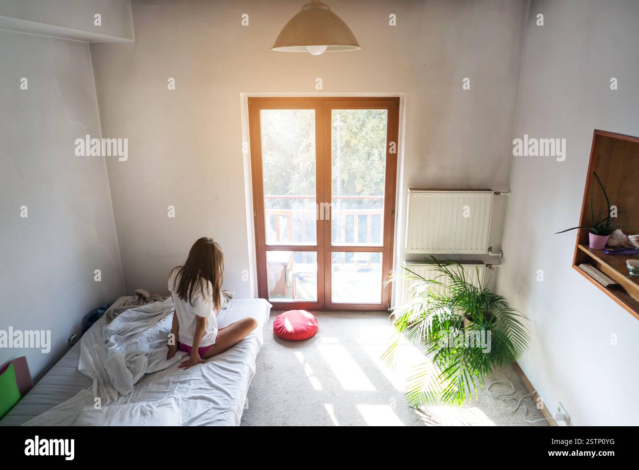 Young woman sitting on bed and looking through the window after waking ...