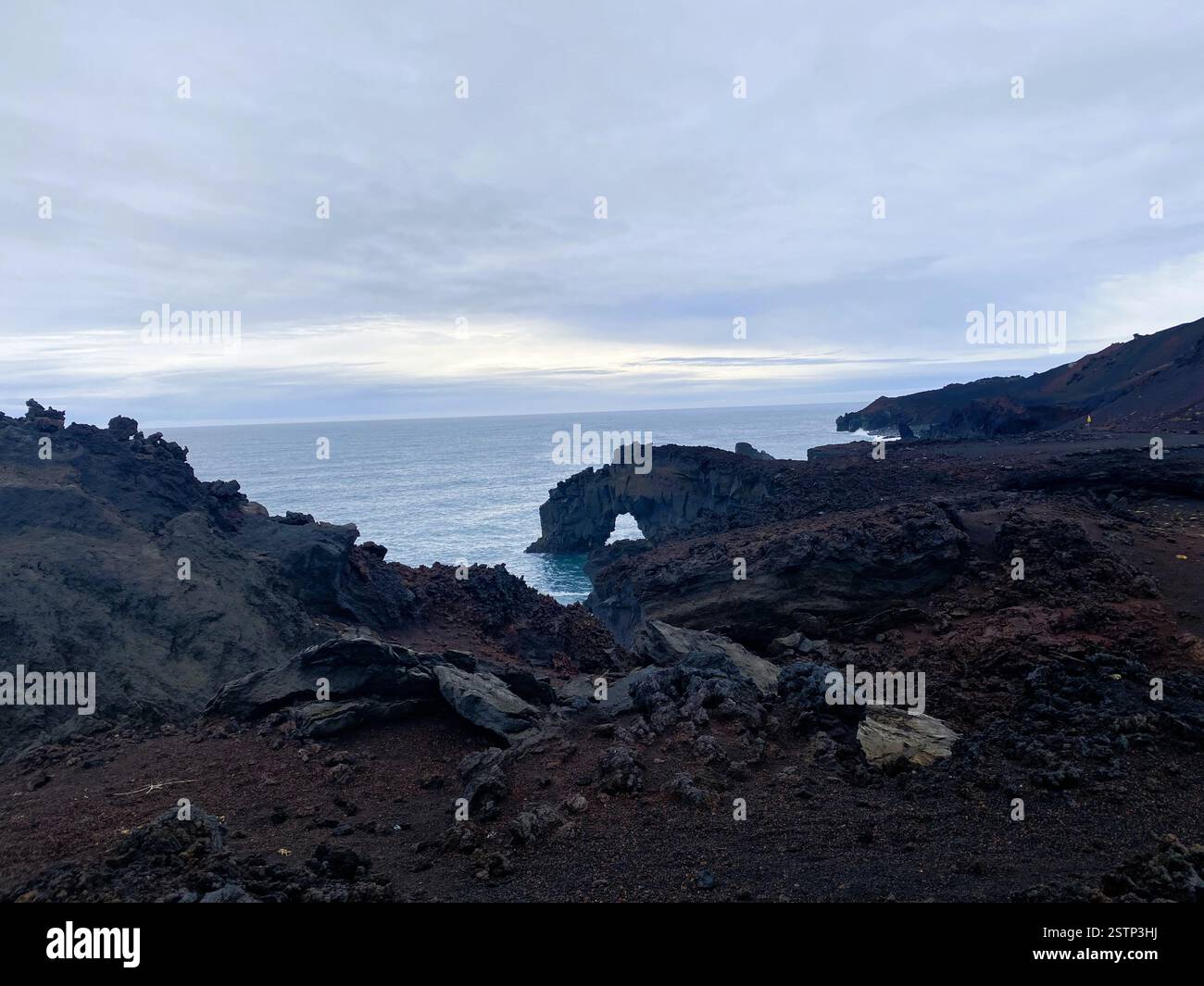 The image depicts a rugged coastal landscape with dark volcanic rocks ...