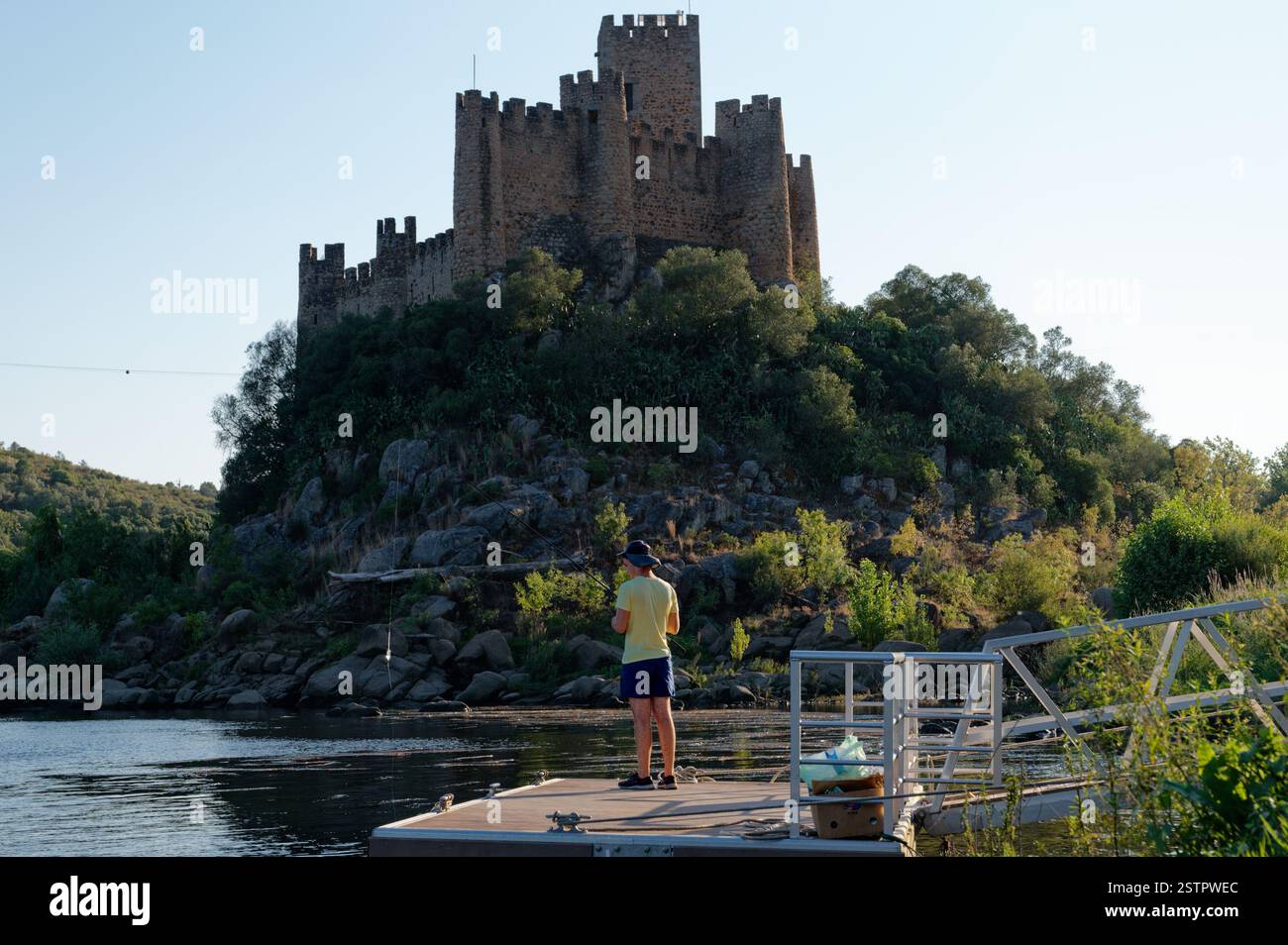 A young man fishes from a small dock with Almourol Castle towering in ...