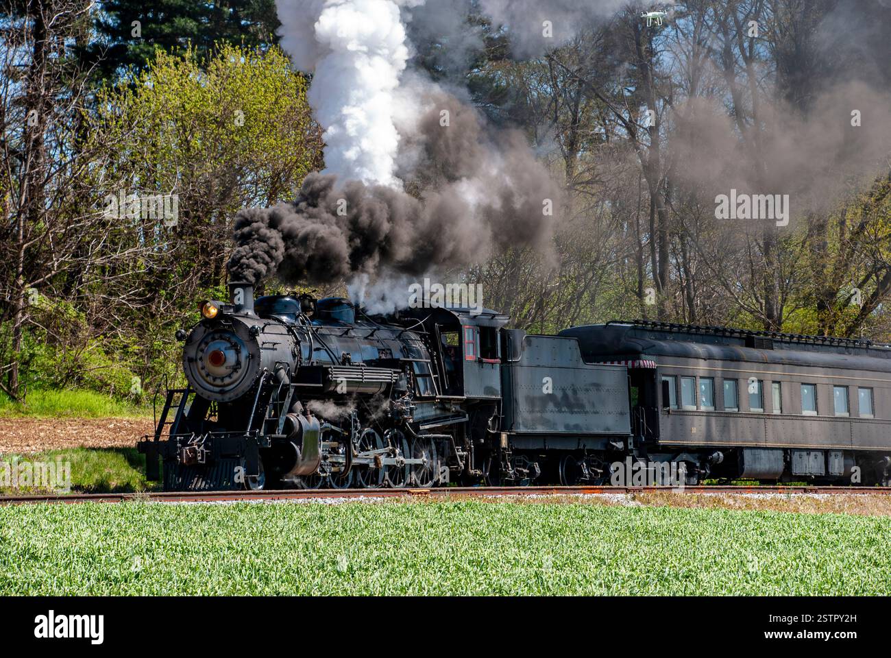 A vintage steam locomotive chugs along a green field, releasing thick ...