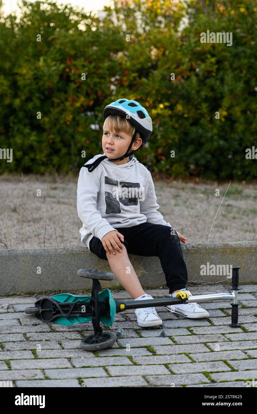 A boy in a helmet sits on the curb, rubbing his knee after falling off ...