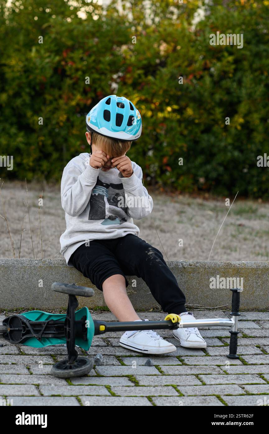 A boy in a helmet sits on the curb, rubbing his knee after falling off ...