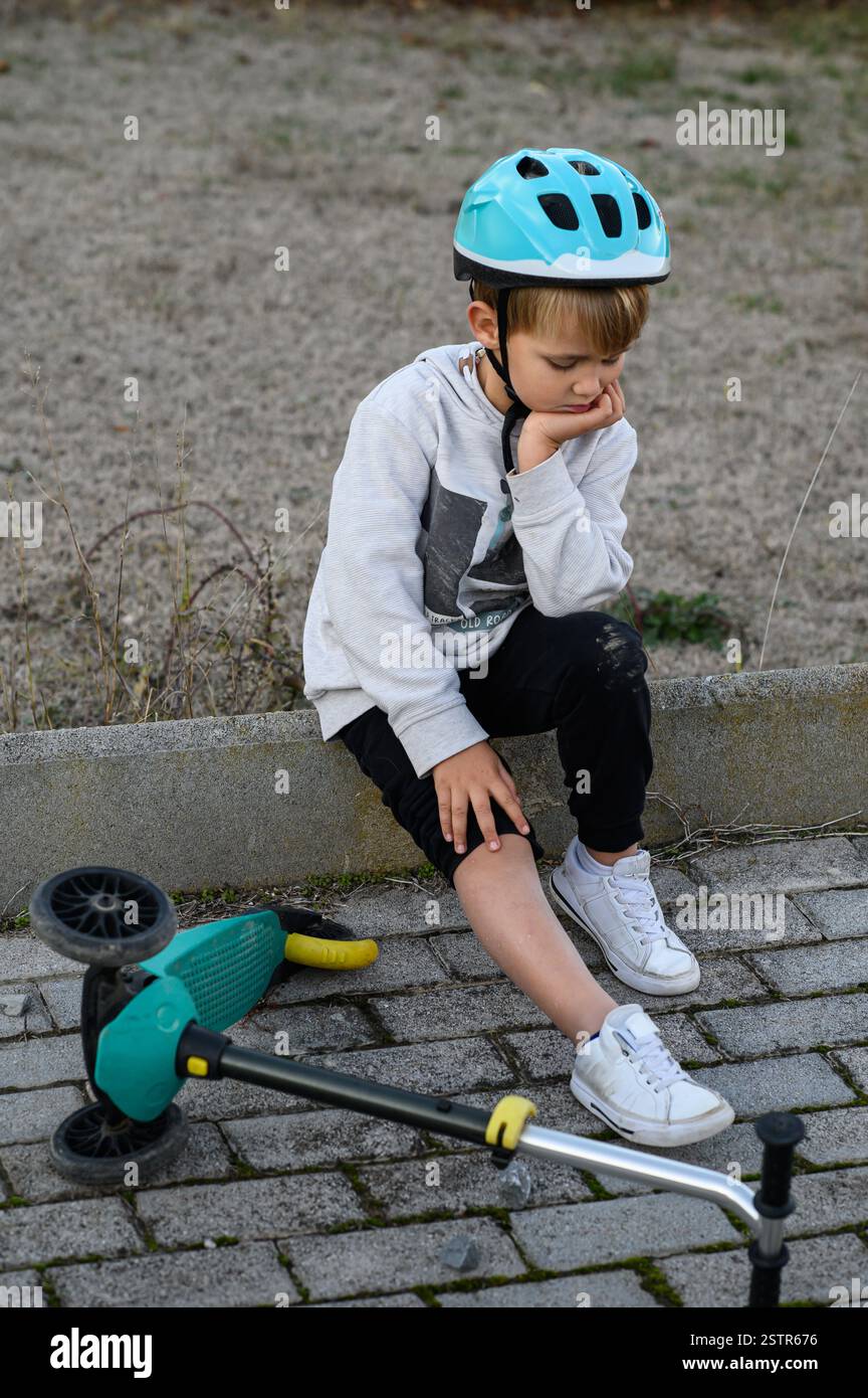 A boy in a helmet sits on the curb, rubbing his knee after falling off ...