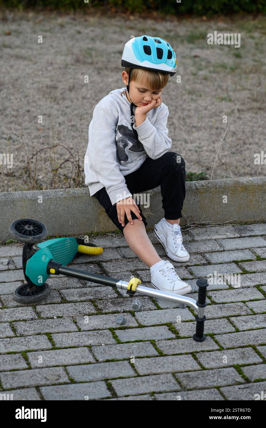 A boy in a helmet sits on the curb, rubbing his knee after falling off ...