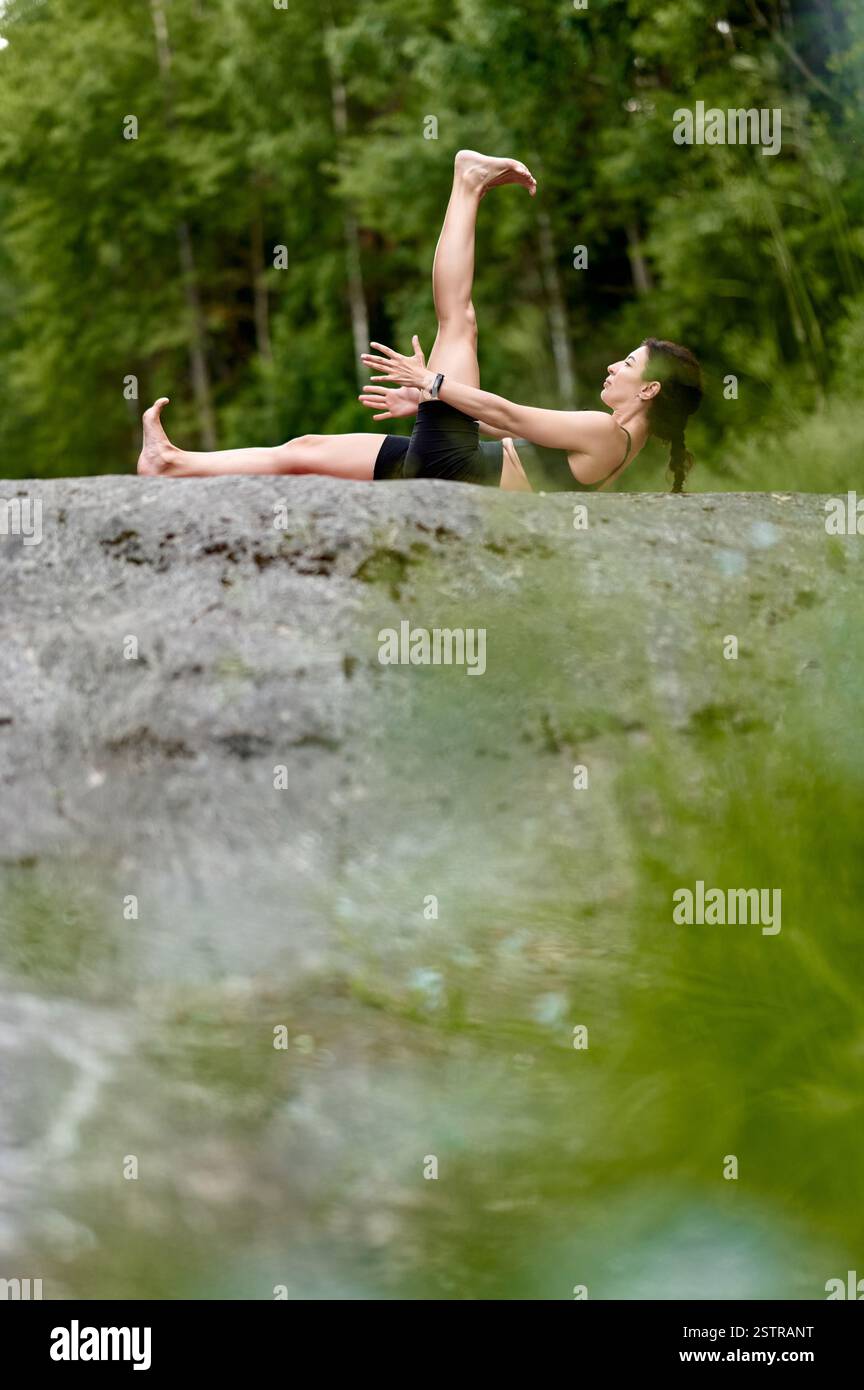 Yoga trainer lying on rock surface, one leg extended straight up, other ...