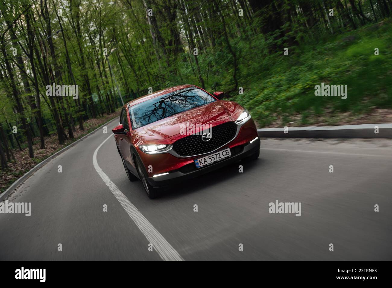 Red Mazda CX-30 on a road in green forest. Front view of compact ...