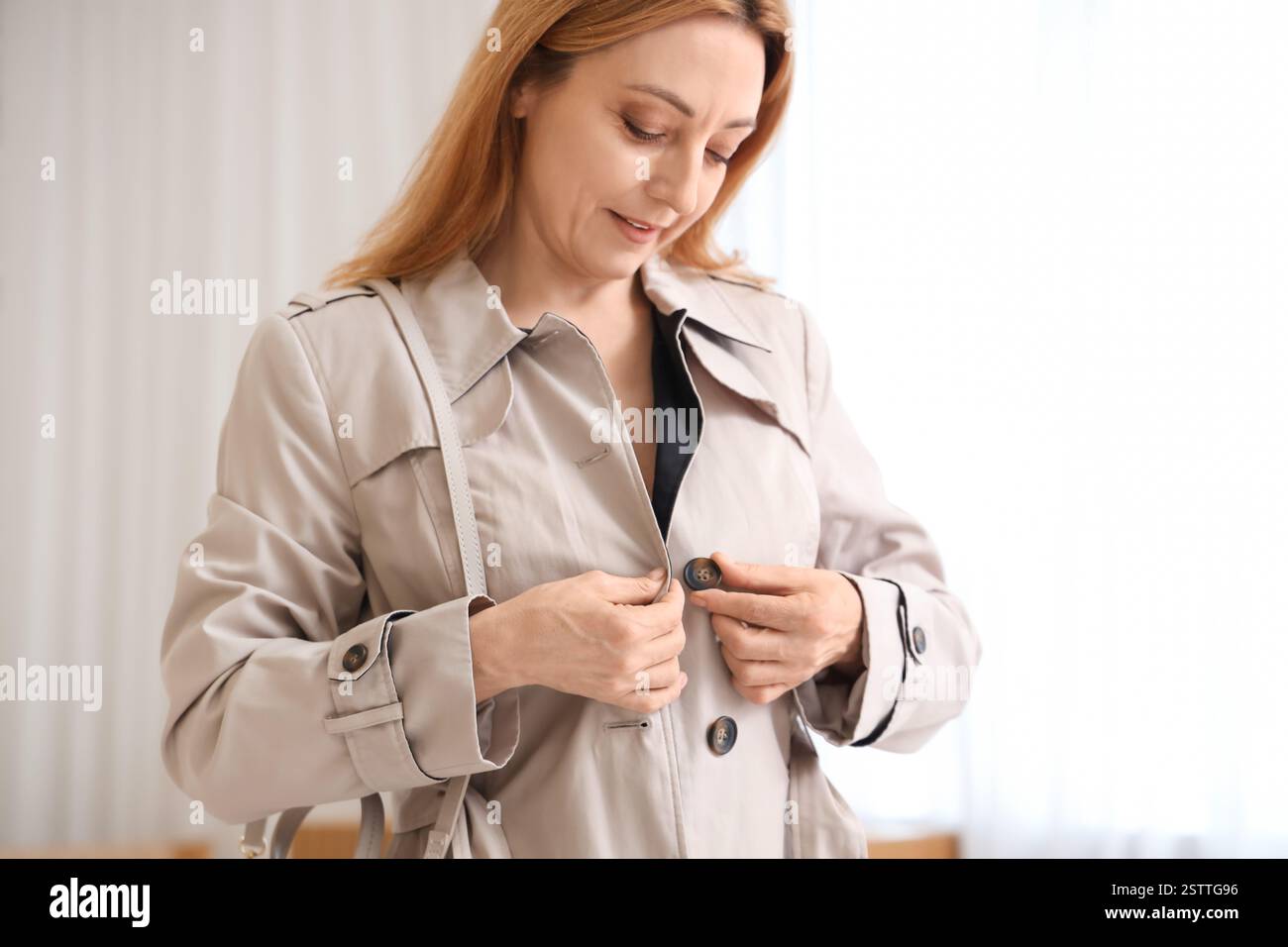 Mature woman buttoning up trench coat at home Stock Photo - Alamy