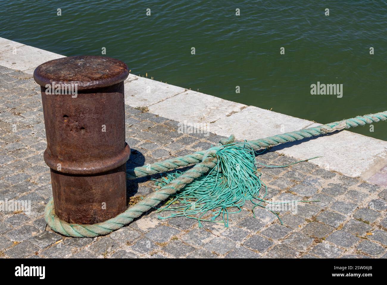 Green mooring rope tied around a rusty metal bollard on a cobblestone ...