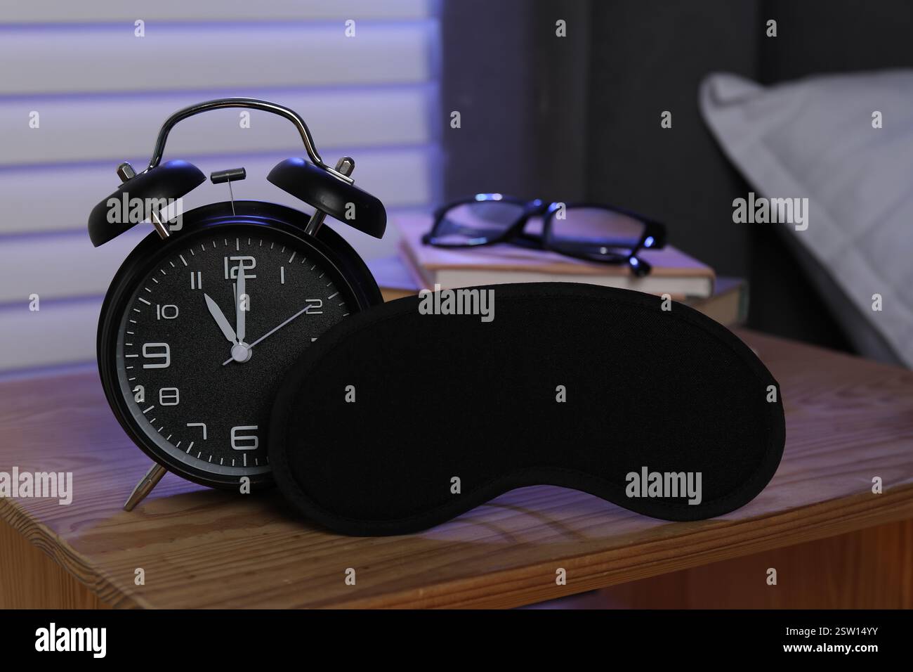 Alarm clock, sleeping mask, books and glasses on nightstand near bed ...