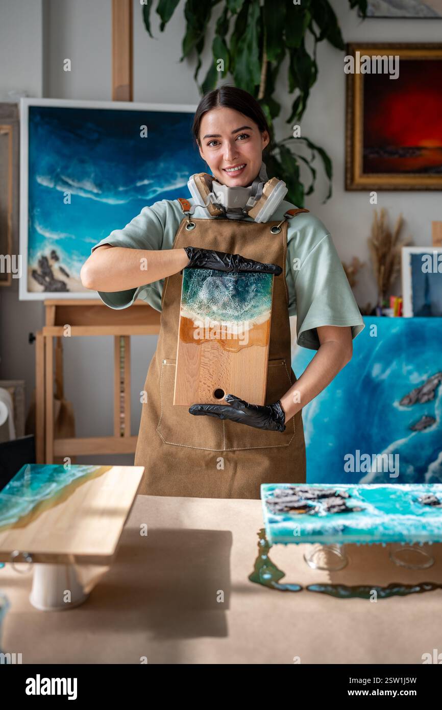 Joyous female artist smiles holding wooden board with completed work in ...