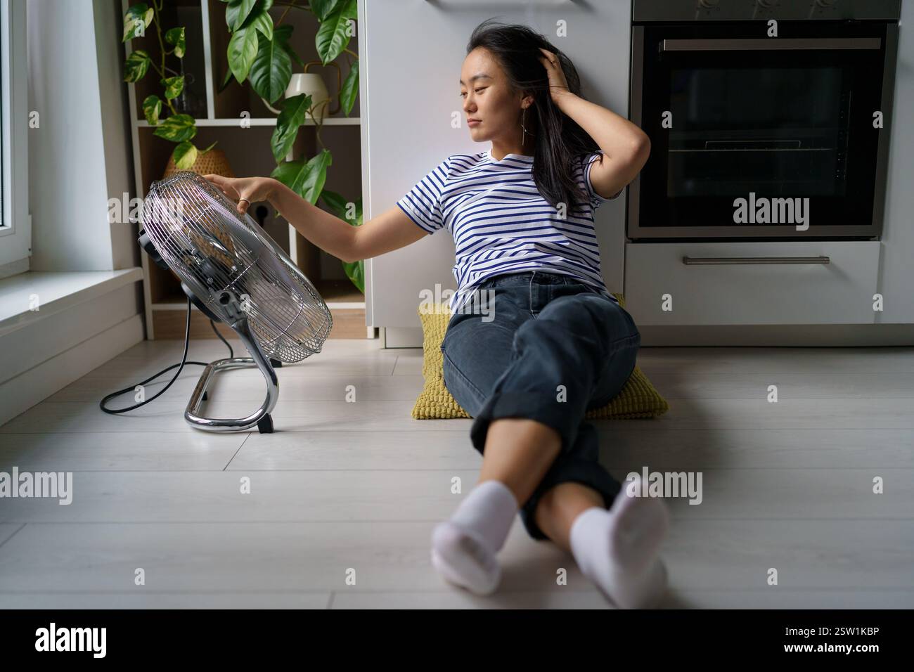 Pleased Asian woman blown by electric fan sits on floor enjoy cool wind ...