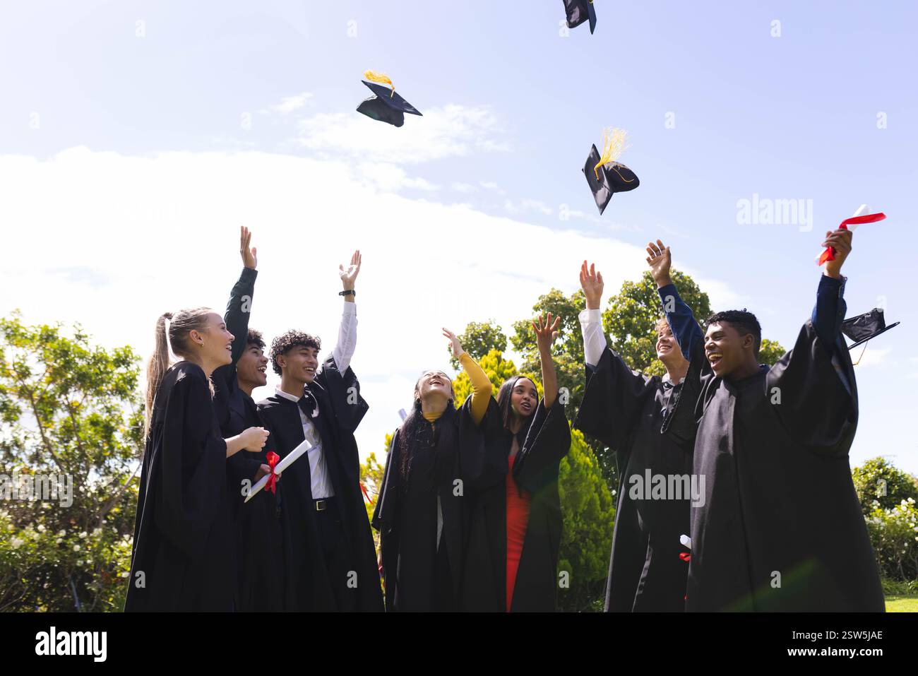 Graduating diverse students joyfully throwing caps in air, celebrating achievement in garden ...