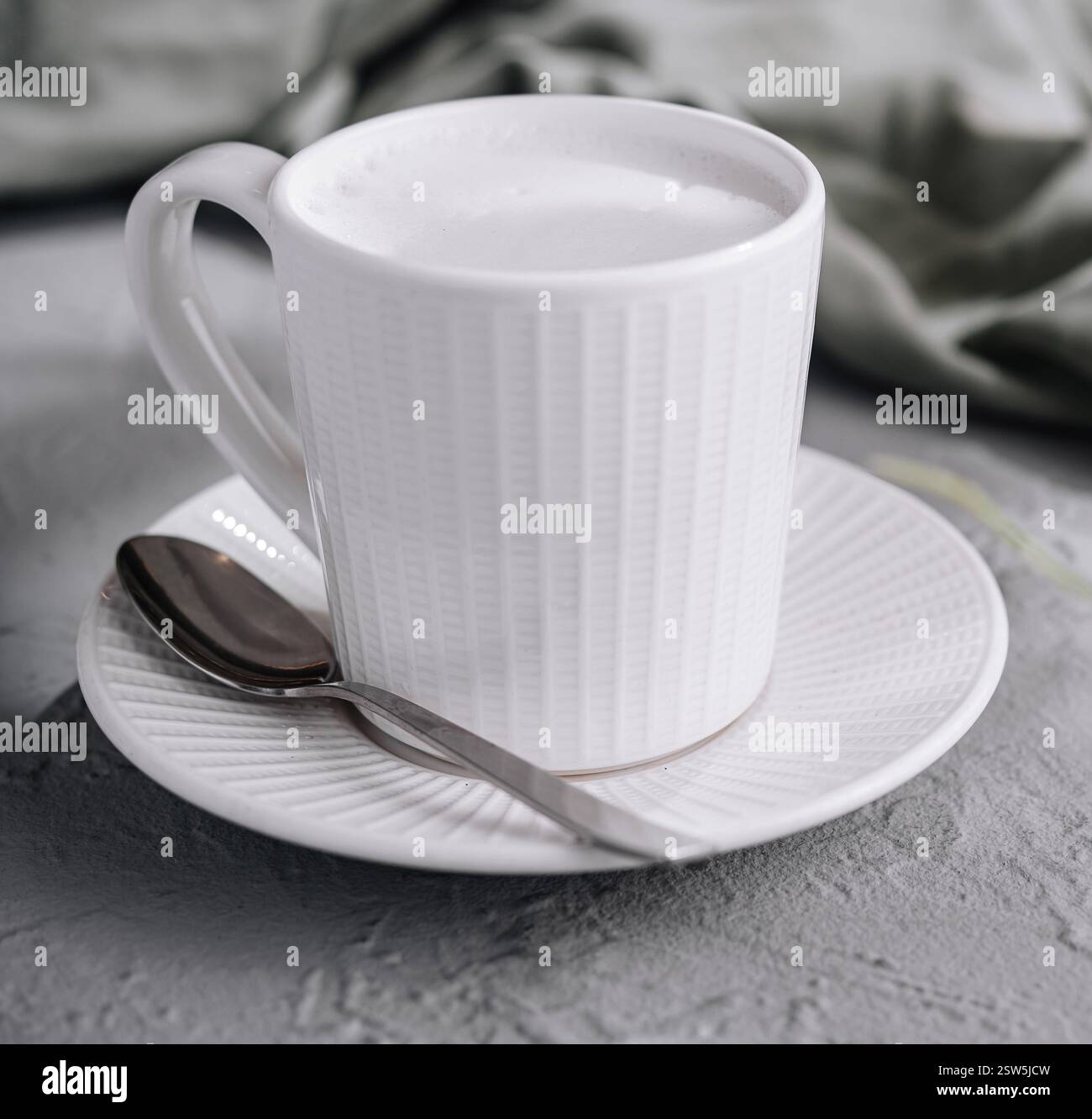 White cup of frothy milk coffee on a textured table with a spoon and ...