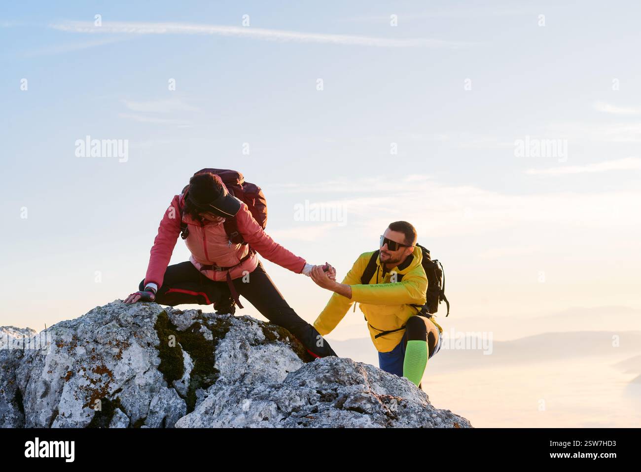 Two hikers, couple in outdoor gear climb rocky terrain with the sun ...