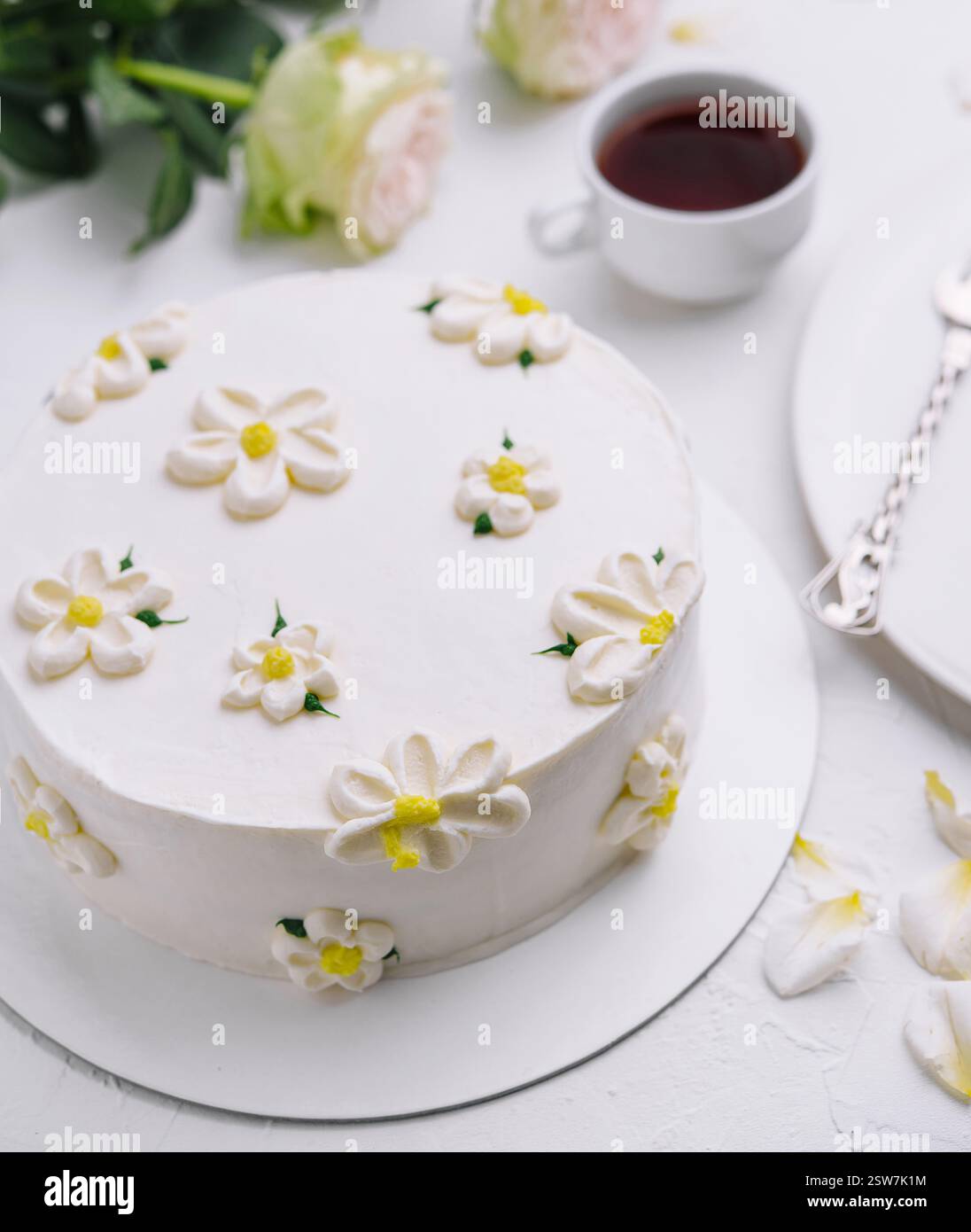 White frosted cake adorned with flowers, alongside tea and roses, on a ...