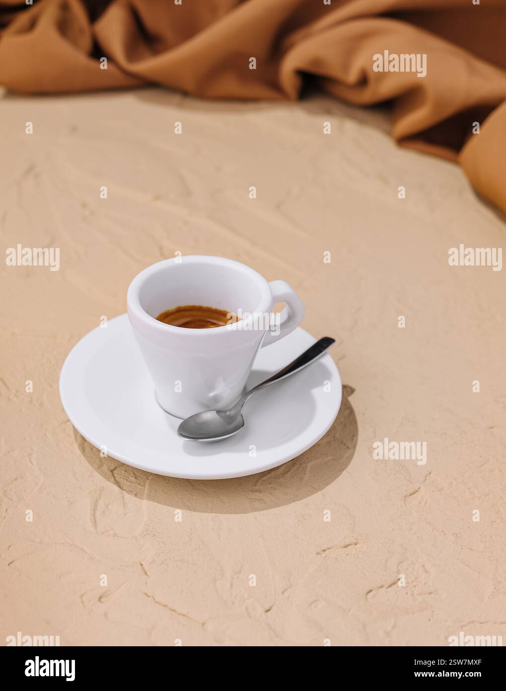 A white cup of espresso coffee on a sandy surface under soft sunlight ...