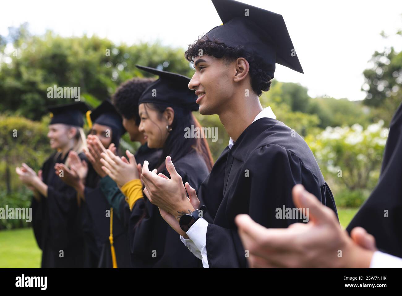 Graduating diverse students in caps and gowns clapping in garden ...