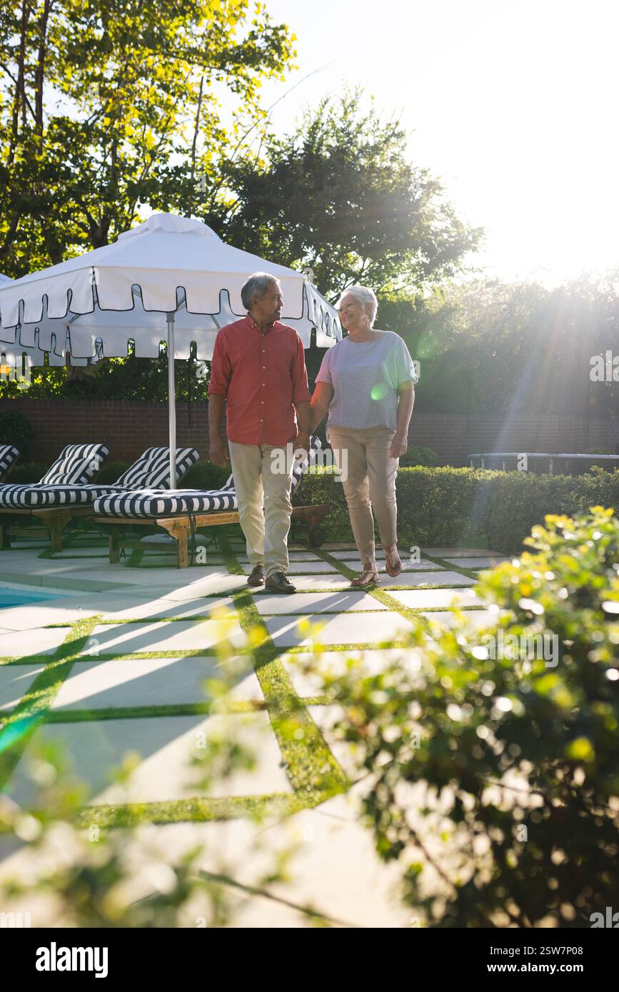 Elderly diverse couple walking hand in hand by poolside, enjoying sunny ...