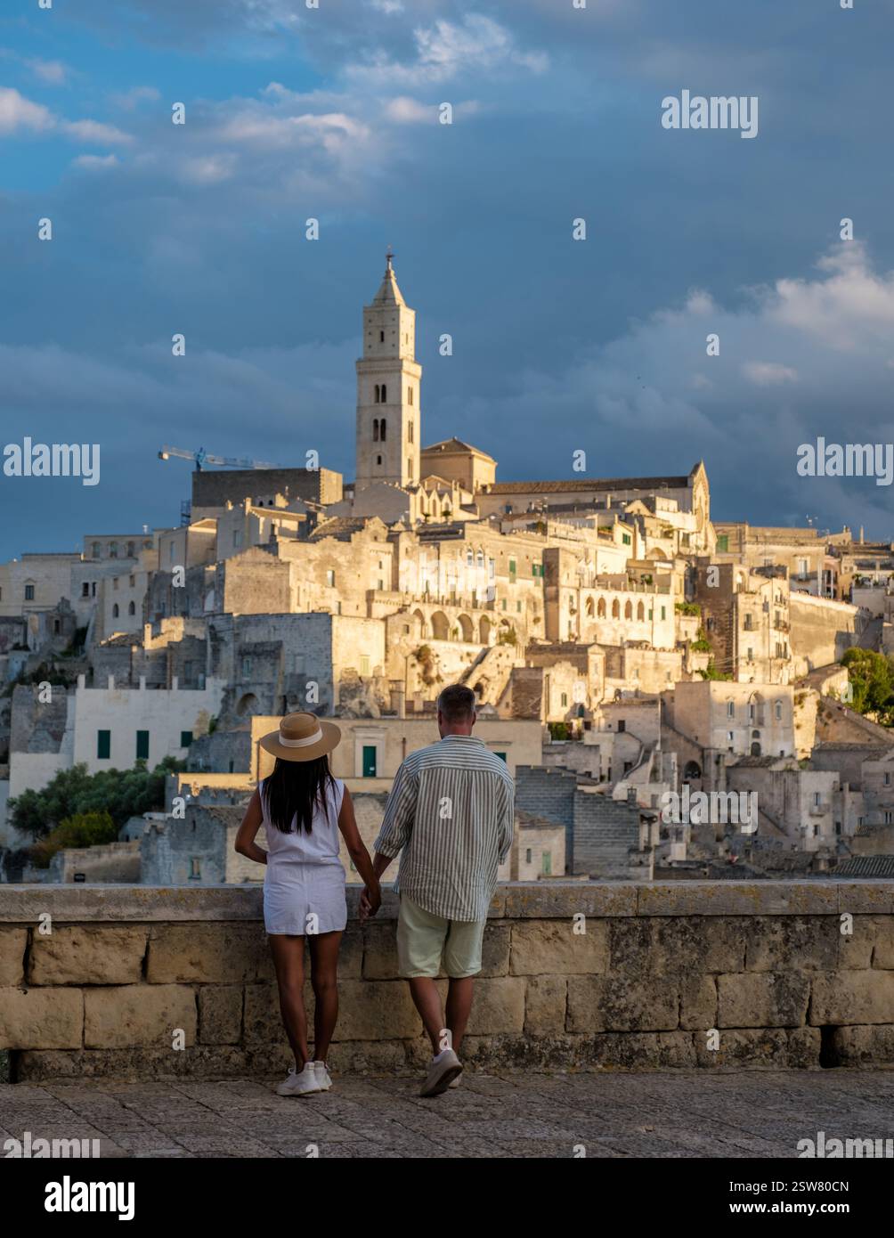 As the sun sets in Matera Italy, a couple holds hands while admiring ...