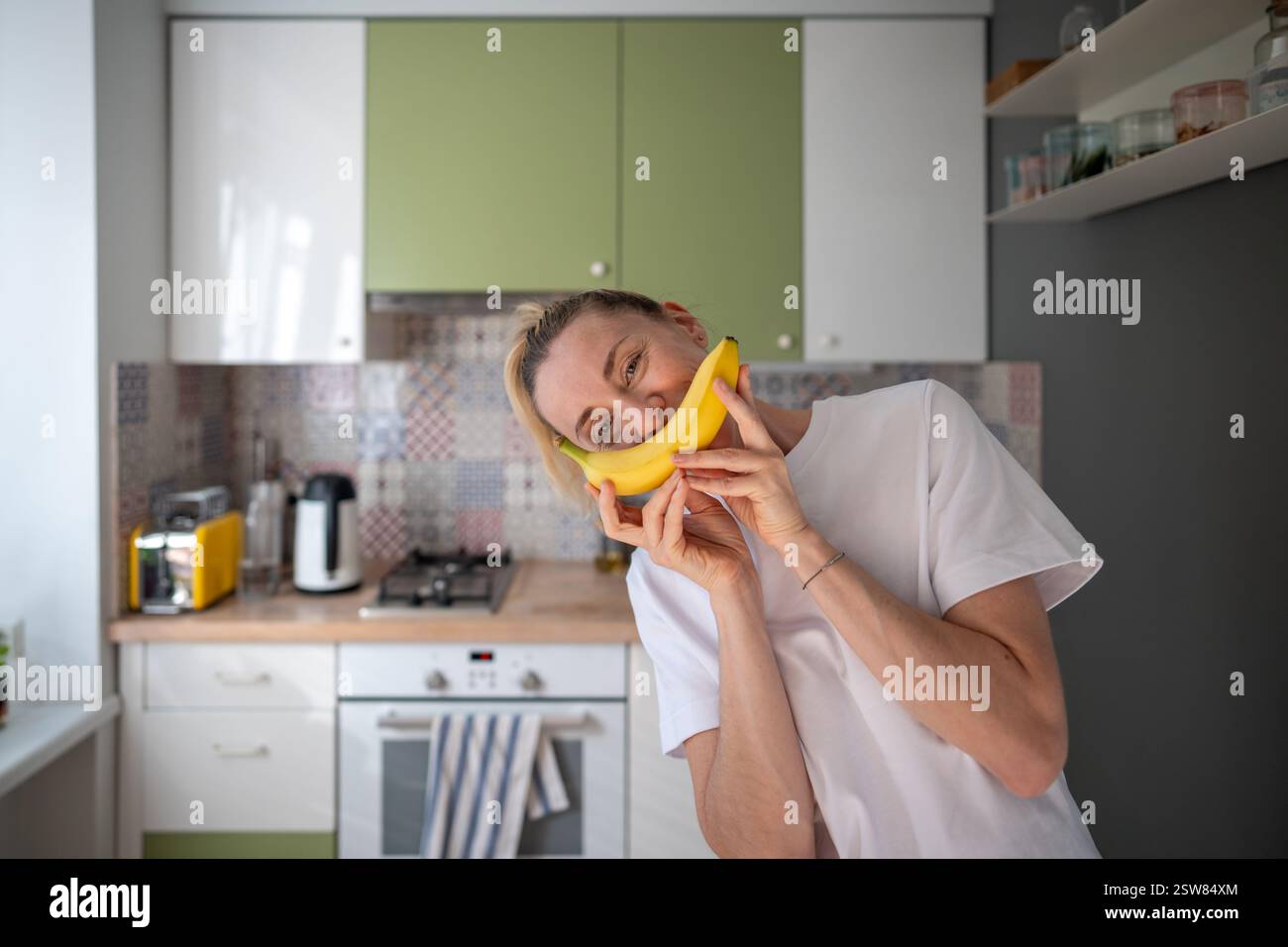 Funny goofy woman puts banana to face like smile in home kitchen ...