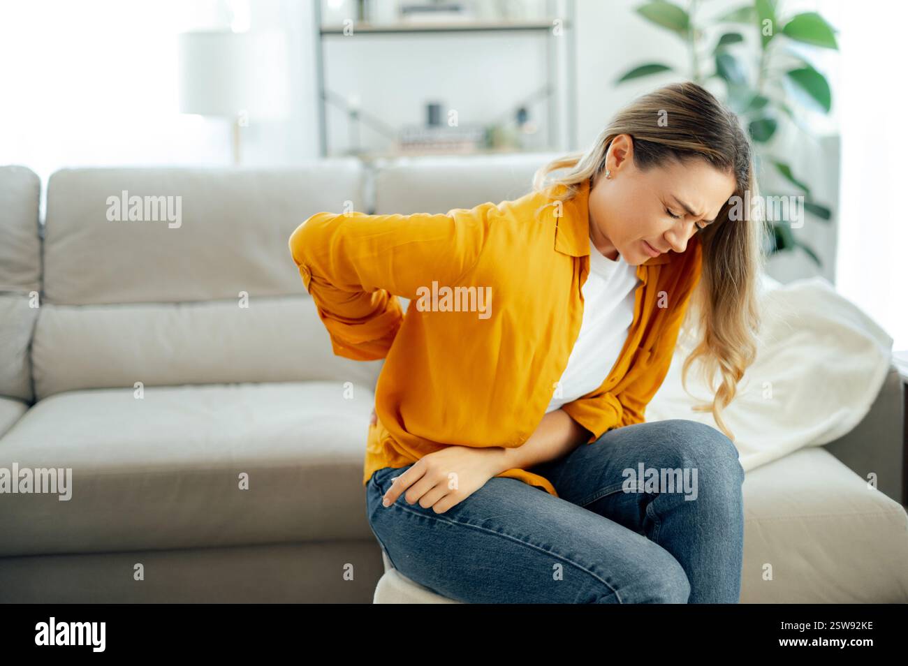 Concept of back pain, discomfort, or muscle strain. Unwell caucasian woman in a yellow shirt ...