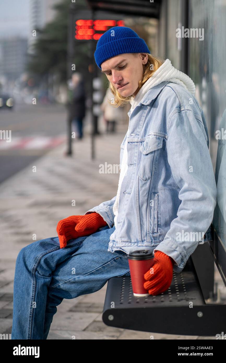 Young man sits on bench at bus stop. Guy with coffee cup calmly waiting ...