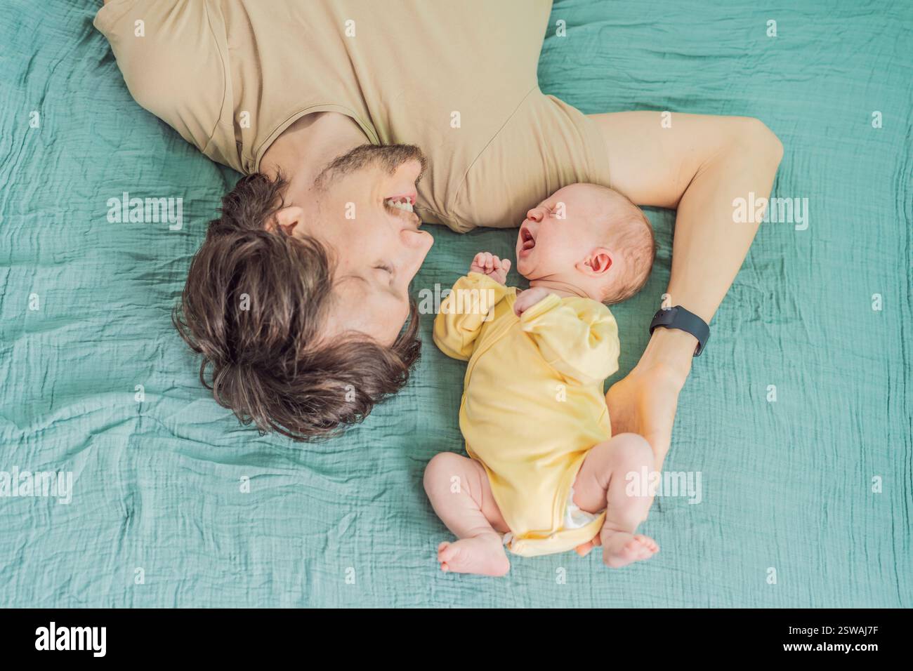 A father and his newborn baby lie on a soft muslin swaddle head to head ...